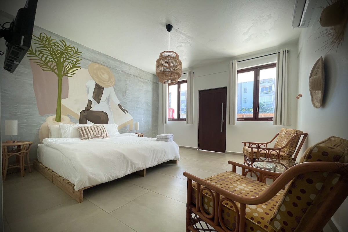 Quarto com uma cama grande, uma cadeira de madeira, uma mesa pequena e um mural na parede.