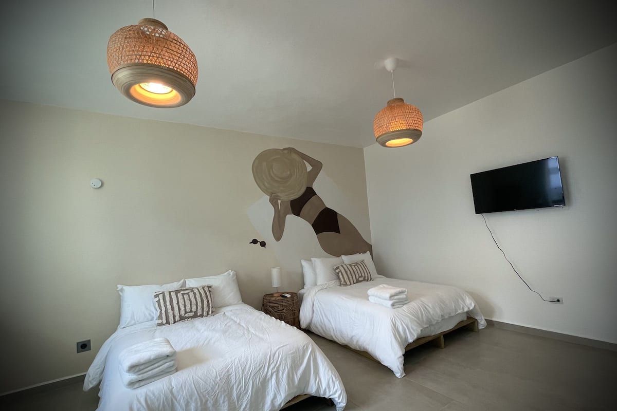 Quarto de hotel com duas camas, televisão de tela plana na parede e duas luminárias pendentes.