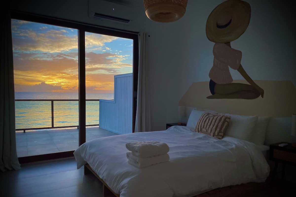 Quarto com cama, mesa de cabeceira, luminária e vista para o oceano ao pôr do sol.