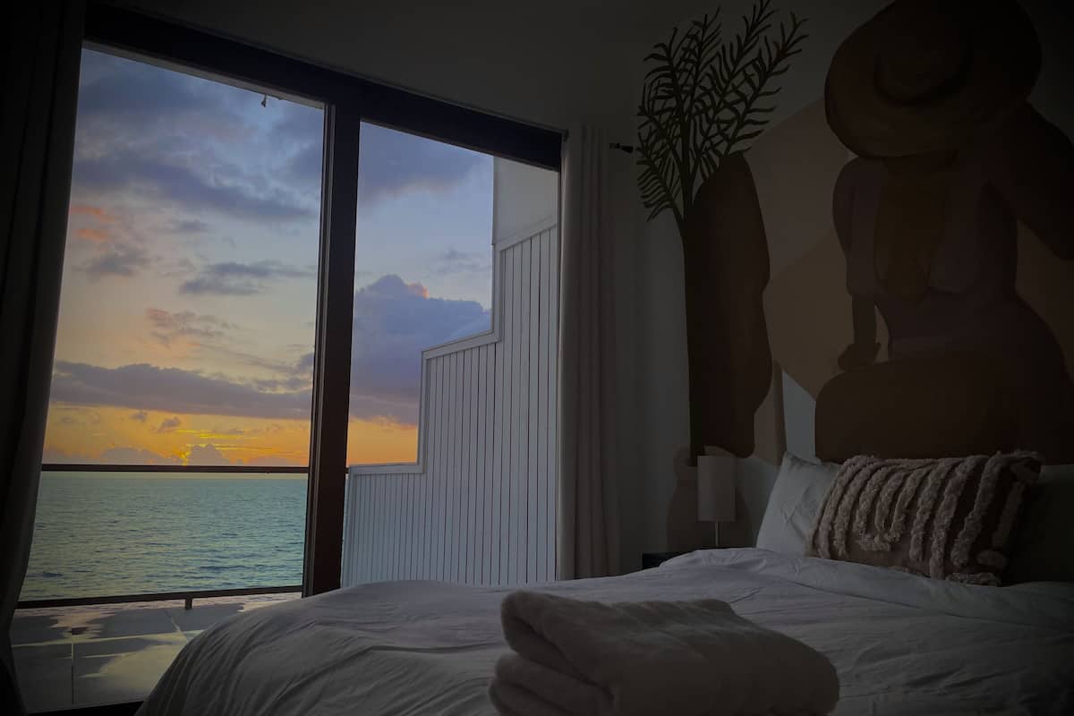 Quarto de hotel com uma grande janela com vista para a água ao pôr do sol.