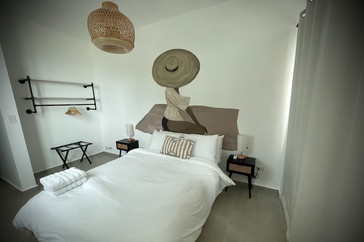 Quarto com uma cama grande, duas mesas de cabeceira, um candeeiro pendente e um porta-toalhas na parede.