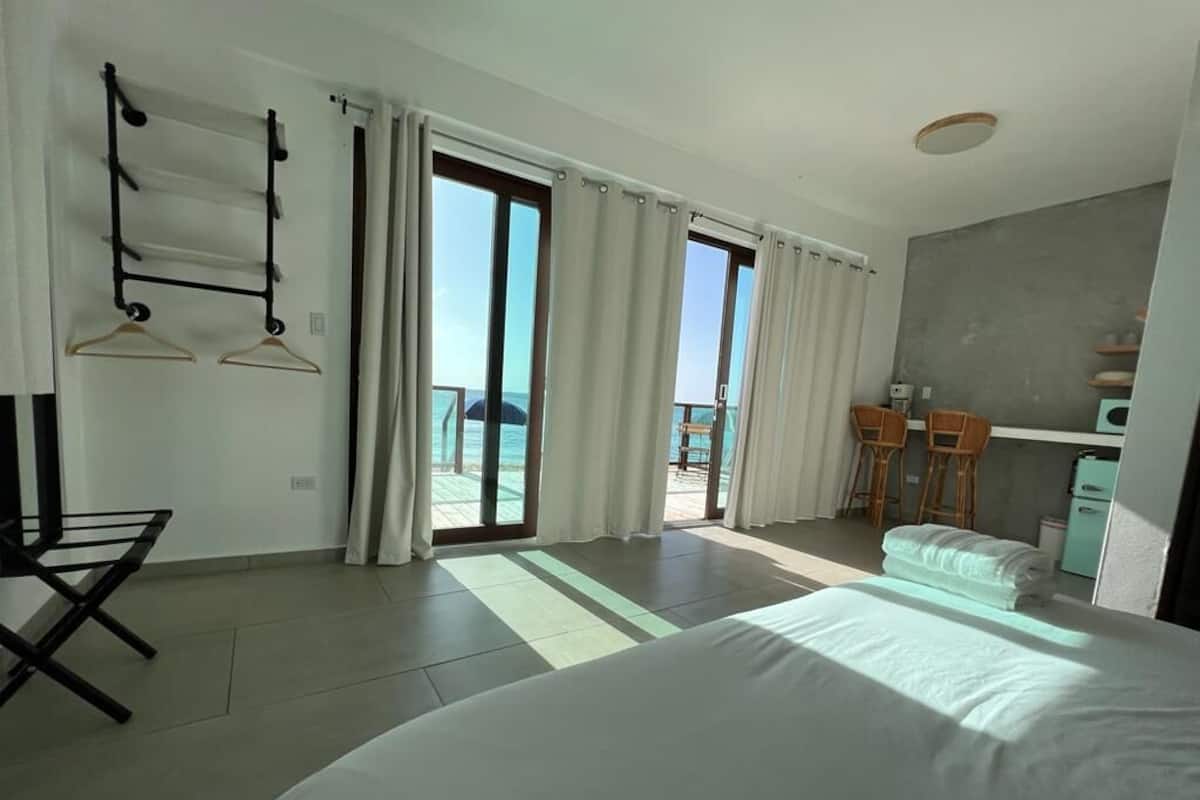 Quarto de hotel com uma cama grande, área de estar com mesa e cadeiras, vista para o mar pelas janelas e varanda com uma cadeira.