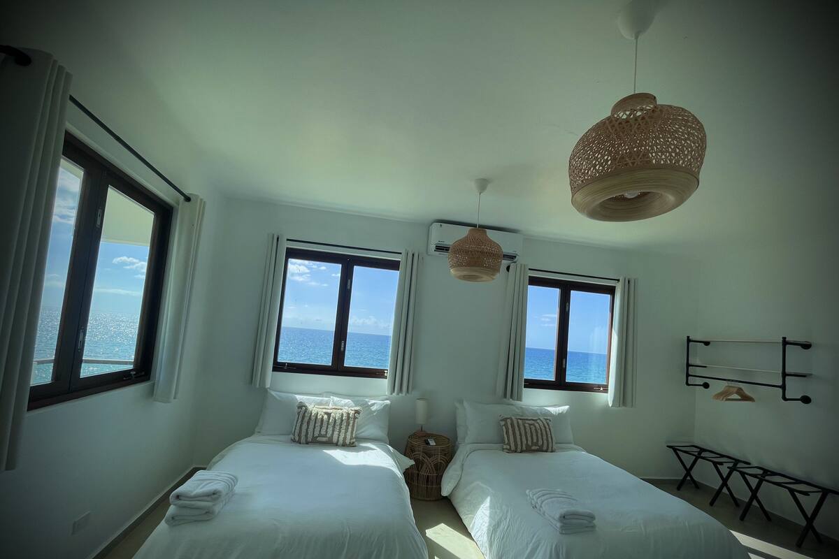 Quarto com duas camas, janelas amplas com vista para o mar, luminária pendente e varal fixado na parede.