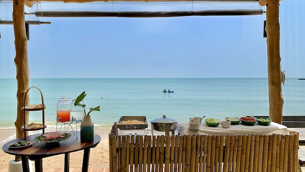 Uma área de refeições à beira-mar com vista para o oceano, uma churrasqueira e diversos alimentos e bebidas sobre uma mesa.