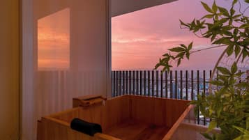 Uma banheira de madeira em um quarto com uma grande janela com vista para a paisagem urbana ao pôr do sol.