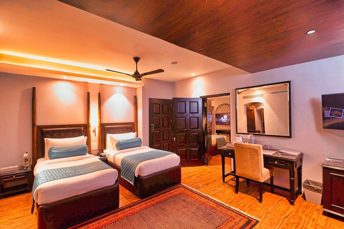 Quarto de hotel com duas camas, uma escrivaninha, uma cadeira, um ventilador de teto e uma televisão.