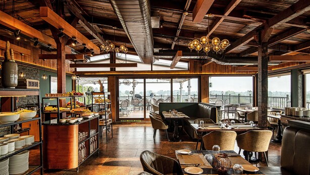 Um restaurante com interior rústico, vigas de madeira e vista através de grandes janelas.
