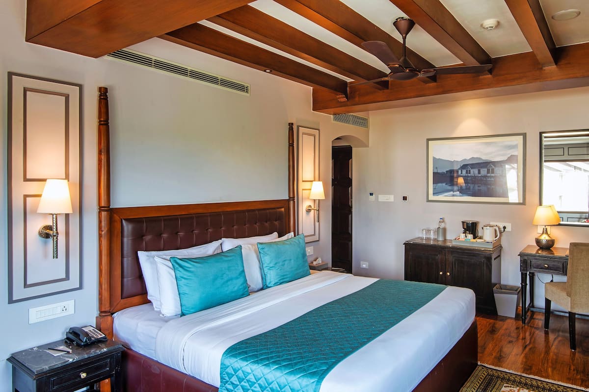 Quarto de hotel com uma cama grande, cabeceira de madeira, duas luminárias de cabeceira, uma escrivaninha com cadeira e um quadro emoldurado na parede.