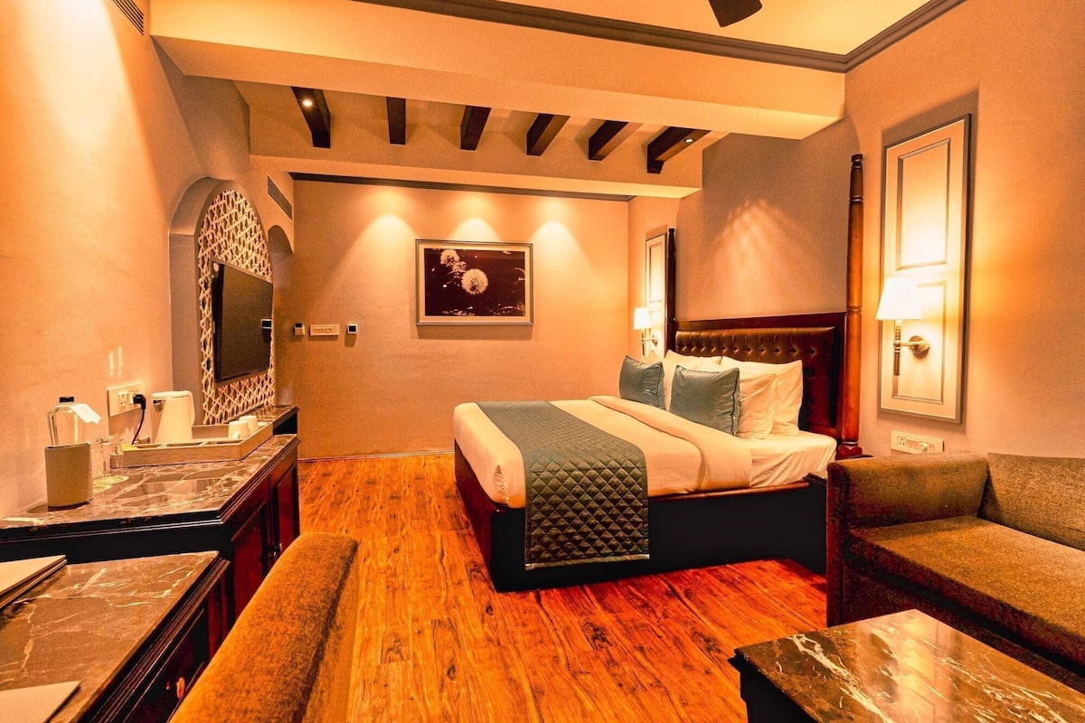 Quarto de hotel com cama, sofá, piso de madeira, mesa de mármore, espelho e quadro na parede.