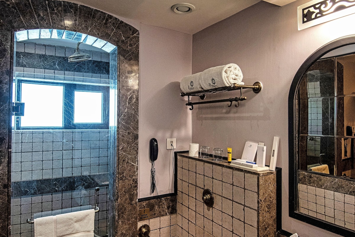 Um banheiro com piso de mármore, vaso sanitário com assento de madeira, pia com torneira dourada, porta-toalhas e área de chuveiro com porta de vidro.