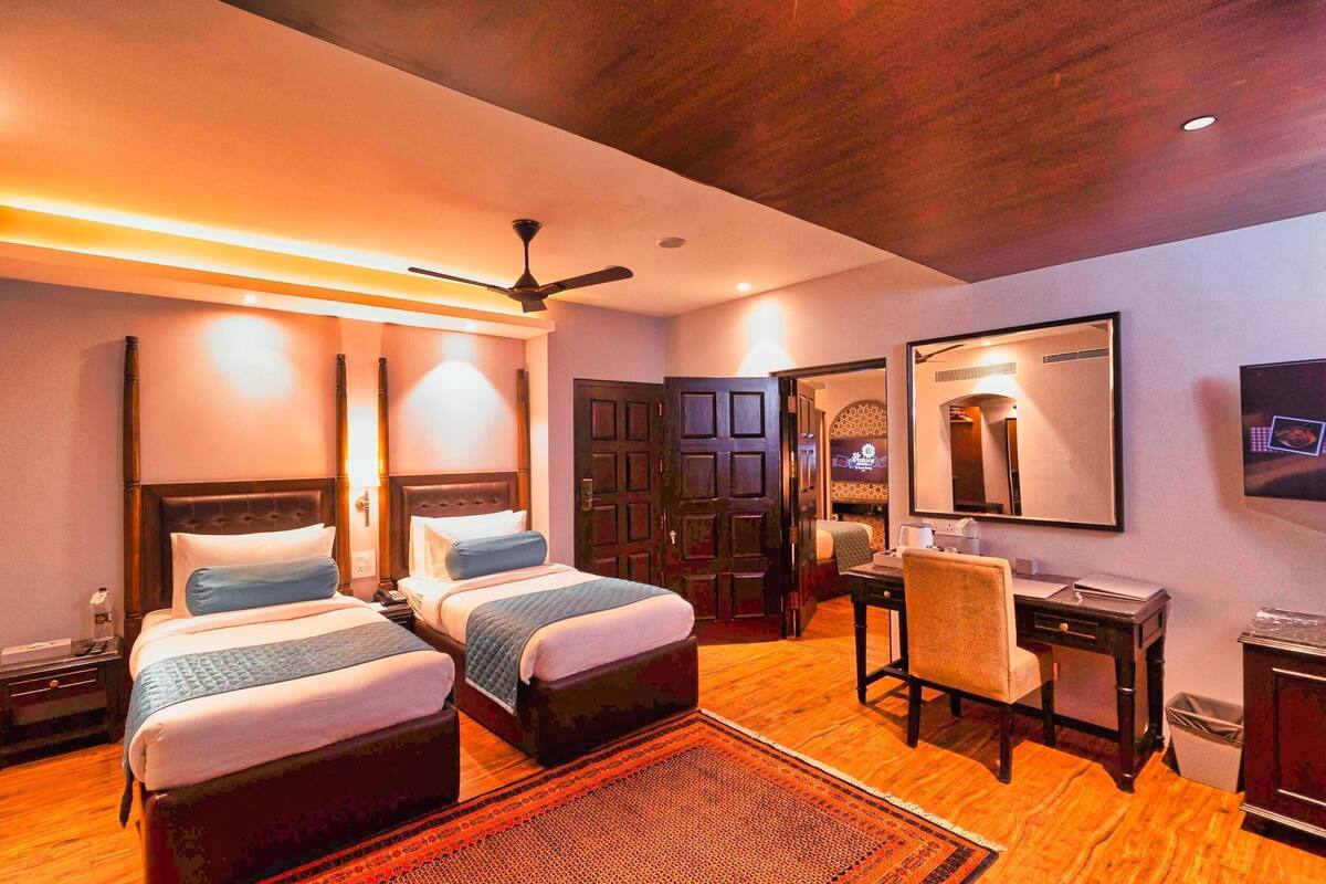 Quarto de hotel com duas camas, uma escrivaninha, uma cadeira, um ventilador de teto e uma televisão.