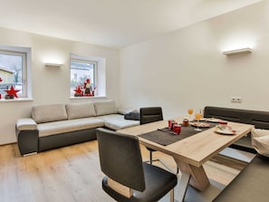 Uma sala de estar moderna com mesa de jantar de madeira, sofá cinza e cadeiras pretas. Há objetos decorativos no parapeito da janela e uma pequena mesa com petiscos e bebidas.