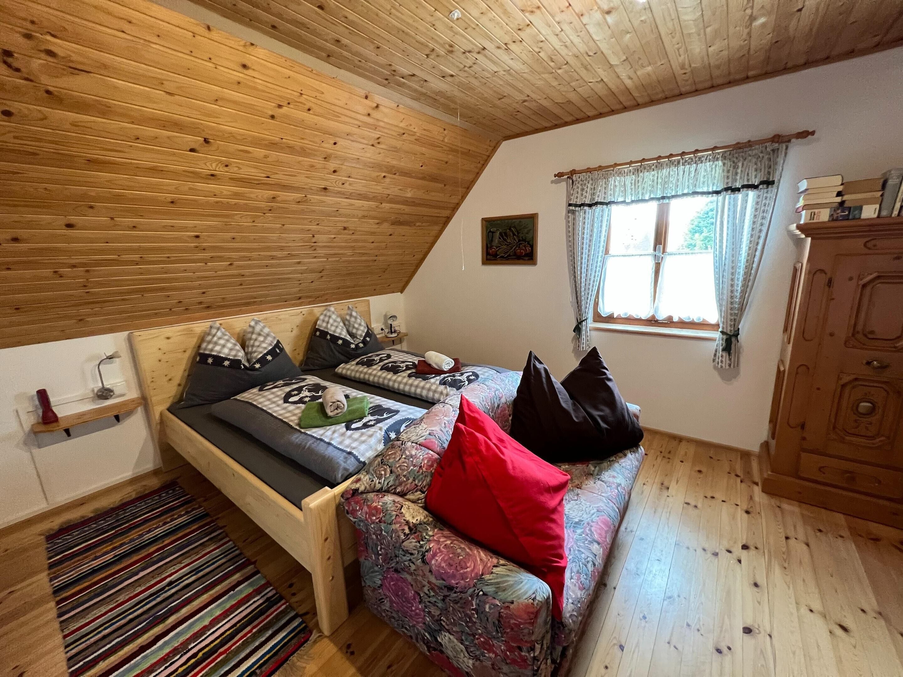 Um quarto acolhedor com teto de madeira, uma cama com roupa de cama estampada, um sofá floral com almofada vermelha e uma janela com cortinas.