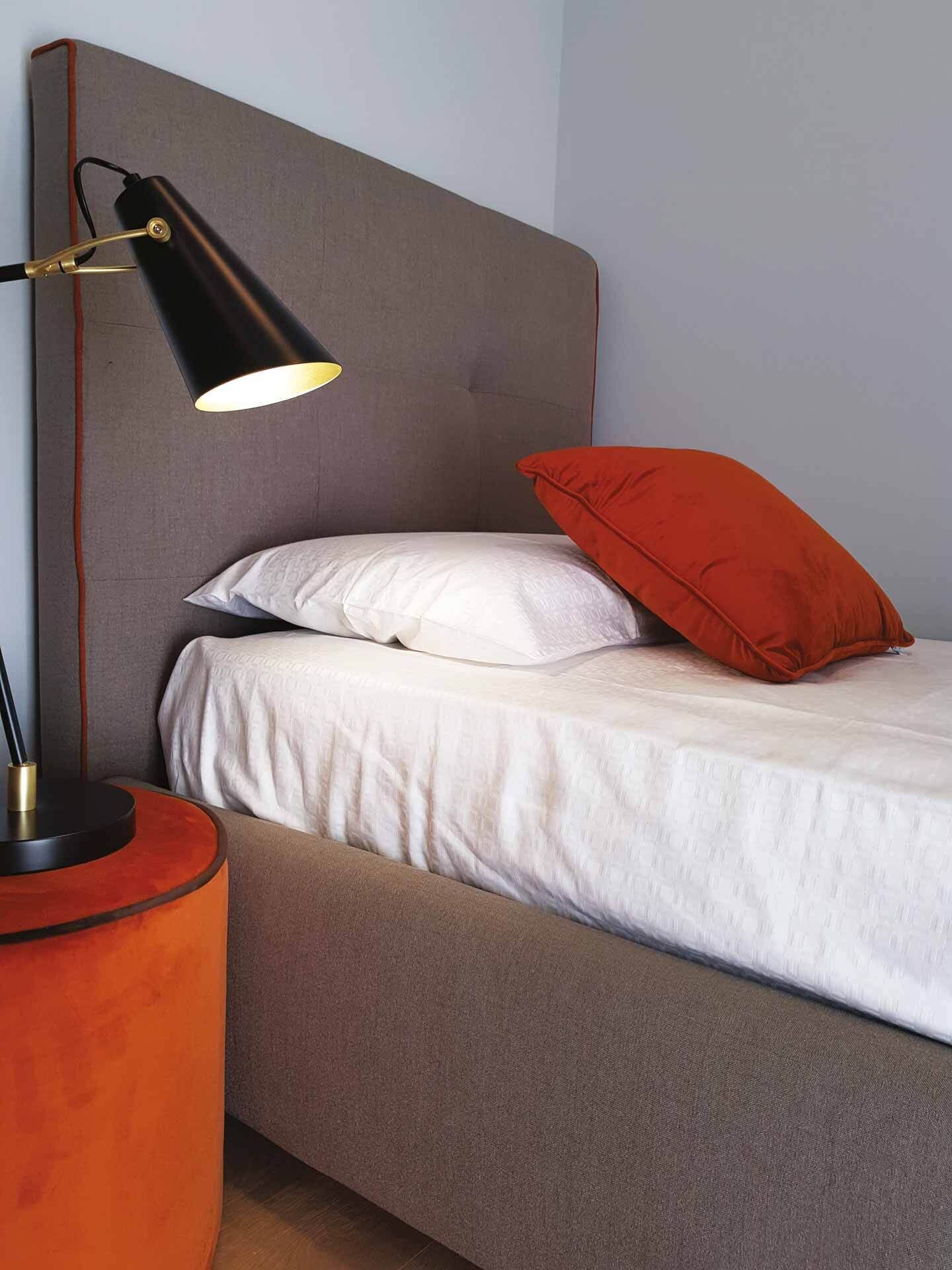 Cama com cabeceira cinzenta, roupa de cama branca e almofada vermelha. Abajur preto com base dourada à esquerda.