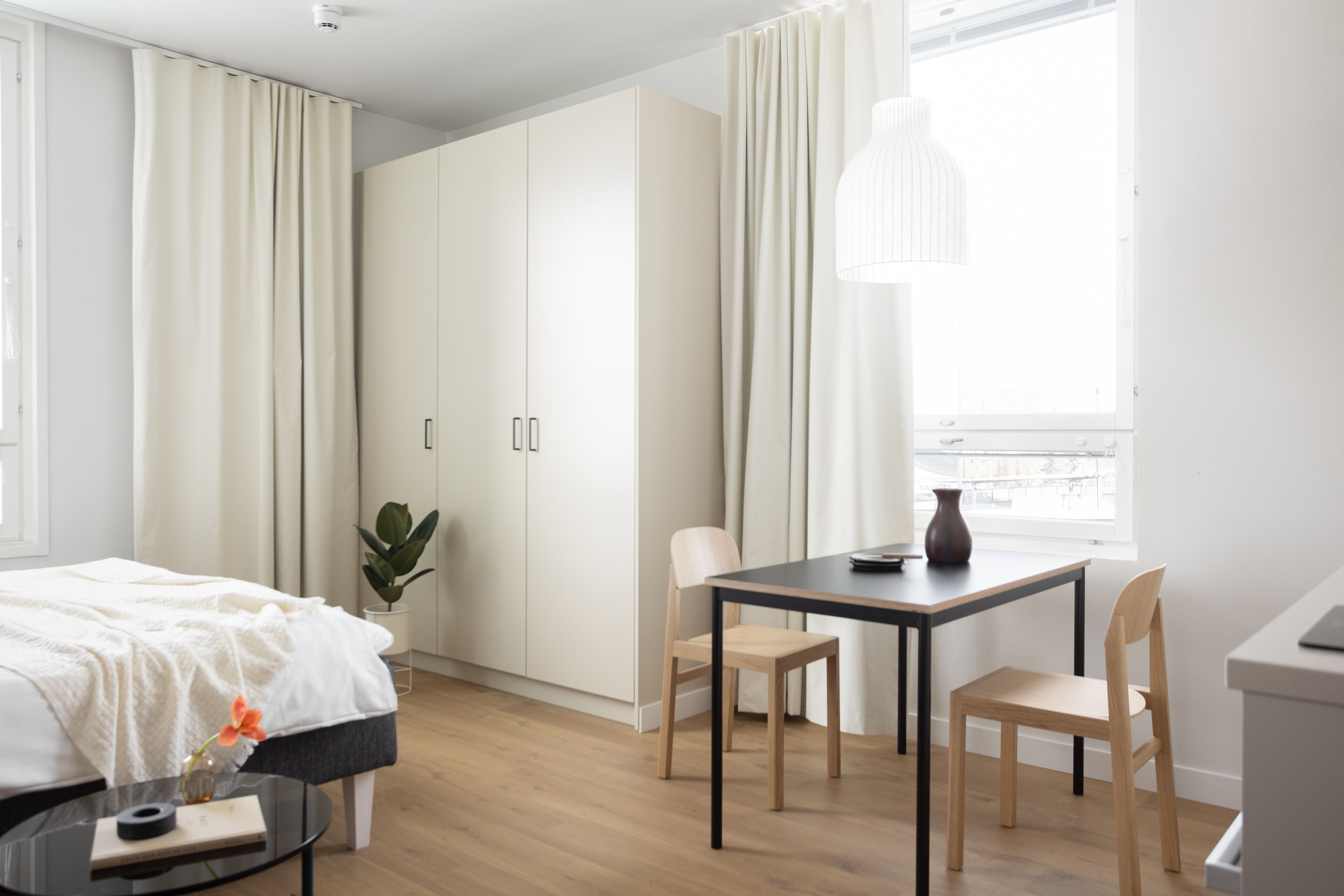 Quarto de hotel moderno com cama, uma mesinha com um vaso, uma cadeira, uma escrivaninha e uma janela com cortinas.