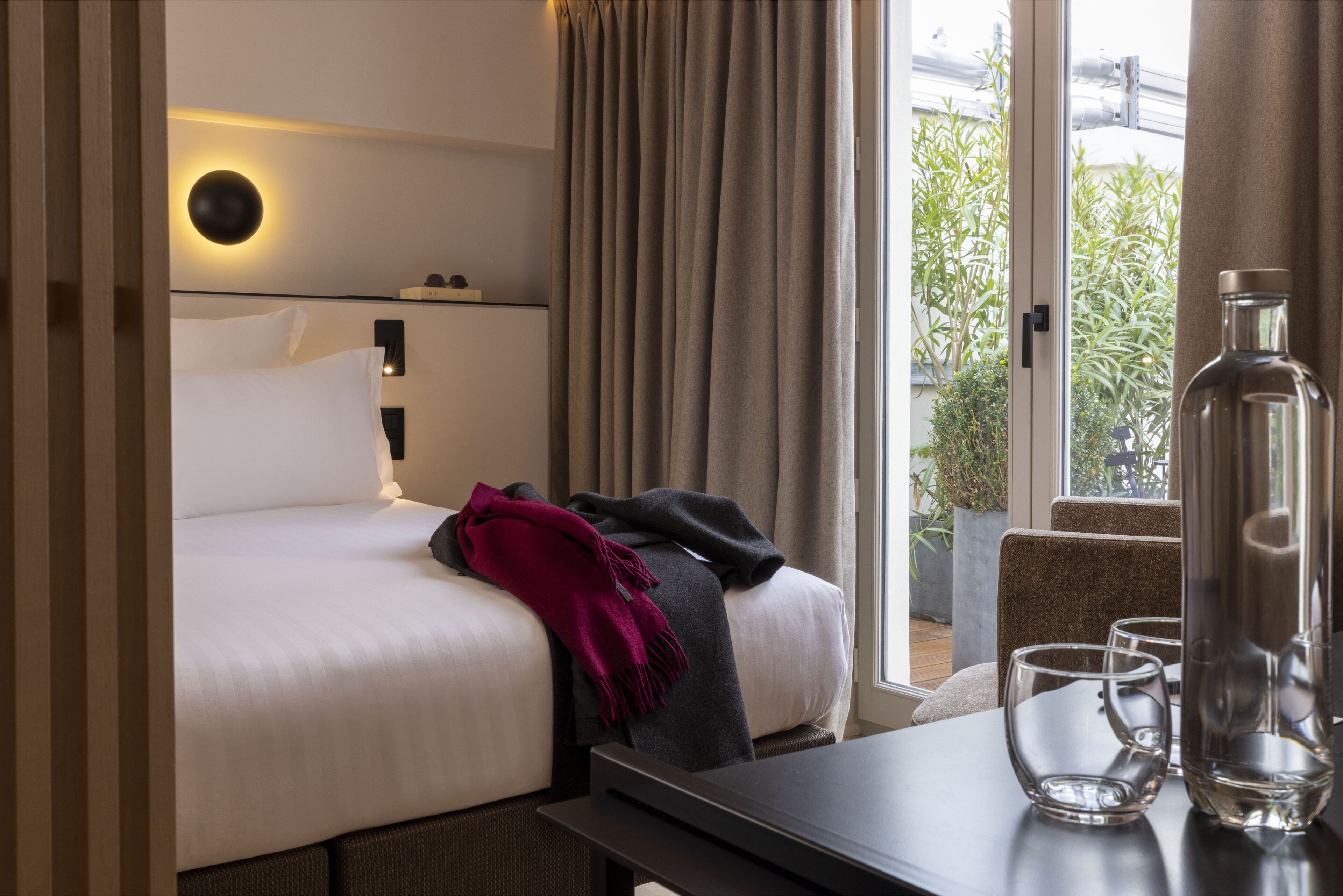 Quarto de hotel com uma cama, uma mesa com copos e uma garrafa, e vista para o verde lá fora.