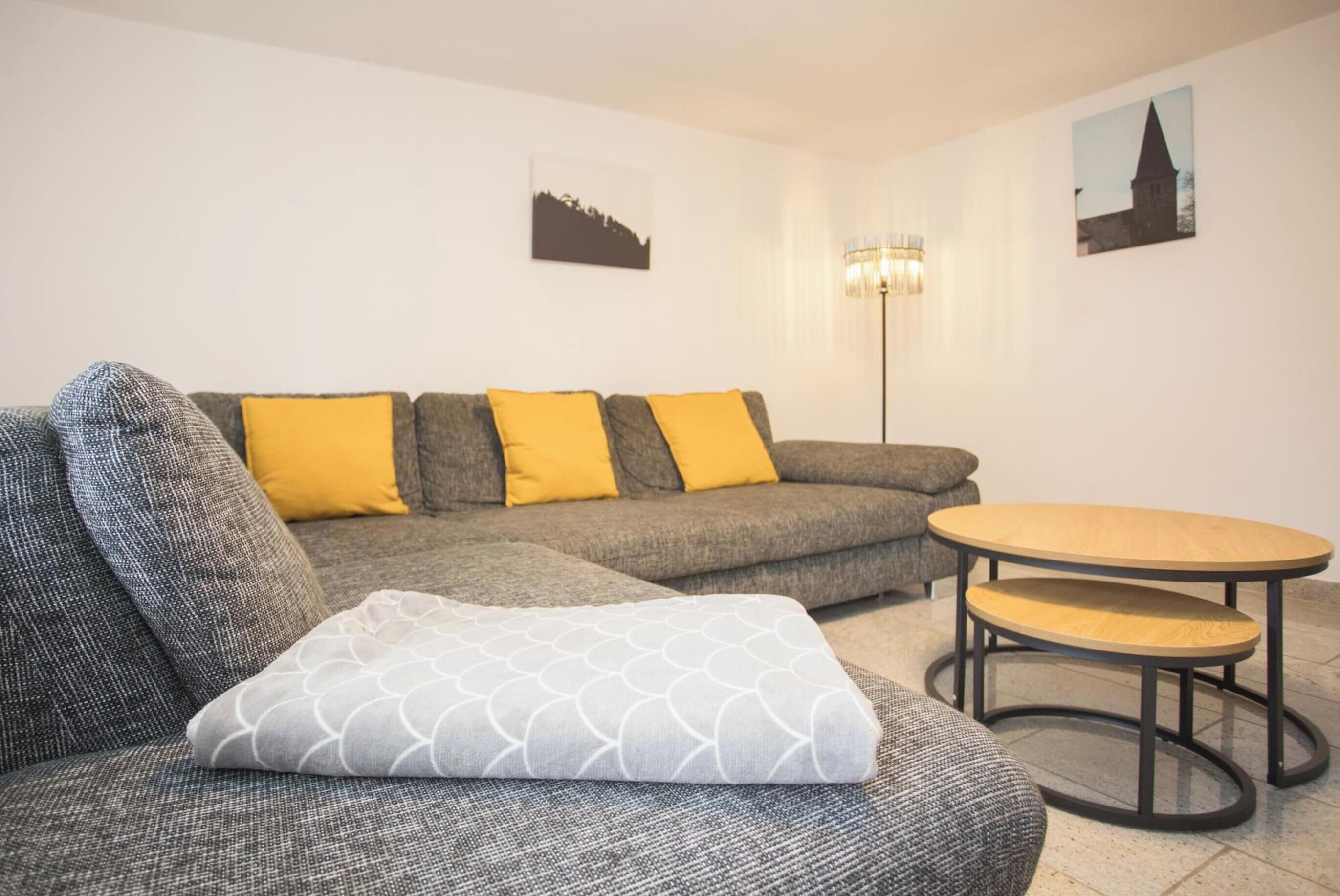 Uma sala de estar moderna com um sofá seccional cinza, duas almofadas amarelas e uma almofada branca com padrão, e uma mesa redonda de madeira com pernas de metal preto.