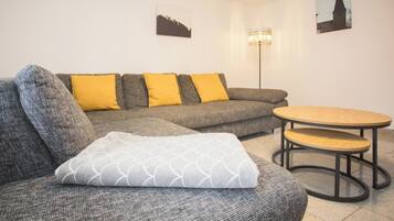 Uma sala de estar moderna com um sofá seccional cinza, duas almofadas amarelas e uma almofada branca com padrão, e uma mesa redonda de madeira com pernas de metal preto.