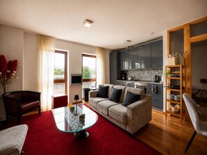 Uma sala de estar moderna com uma mesa de centro de vidro, um sofá, um tapete vermelho e uma cozinha ao fundo.