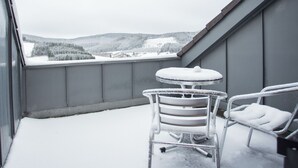 Varanda com mesa de metal e duas cadeiras, coberta de neve, com vista para uma paisagem nevada com árvores e colinas.