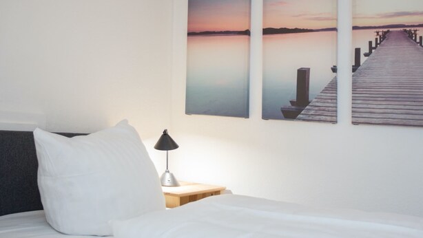 Cama bem arrumada com lençóis e almofadas brancos, uma luminária de cabeceira e uma parede com um grande quadro de paisagem mostrando um cais e o pôr do sol.