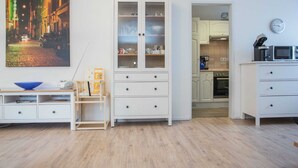 Sala de estar moderna com um armário branco, área de jantar com uma tigela azul e uma cadeira, e cozinha visível por uma porta aberta.