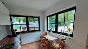 Uma cozinha com área de refeições, duas janelas com molduras pretas, uma mesa pequena com cadeiras e um vaso com planta no parapeito da janela.