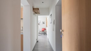 Um corredor com uma porta de madeira à direita, uma parede branca com um telefone e um chave, e uma cadeira com almofada vermelha ao fundo.