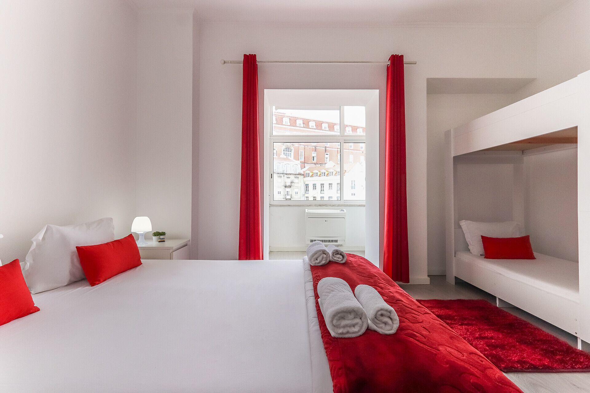 Quarto de hotel com uma cama grande, dois sofás brancos, detalhes vermelhos e vista para edifícios pela janela.