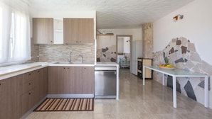 Cozinha moderna com armários de madeira, pia de aço inoxidável e máquina de lavar louça. Apresenta um painel de azulejos e uma parede em mosaico de pedra. Uma pequena mesa com frutas é visível.