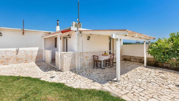 Um alpendre coberto com chão de pedra, uma mesa com cadeiras e um pequeno jardim.