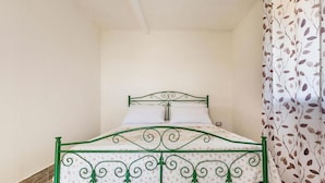 Cama de solteiro com estrutura de metal verde e roupa de cama branca num quarto com paredes lisas e cortina estampada.