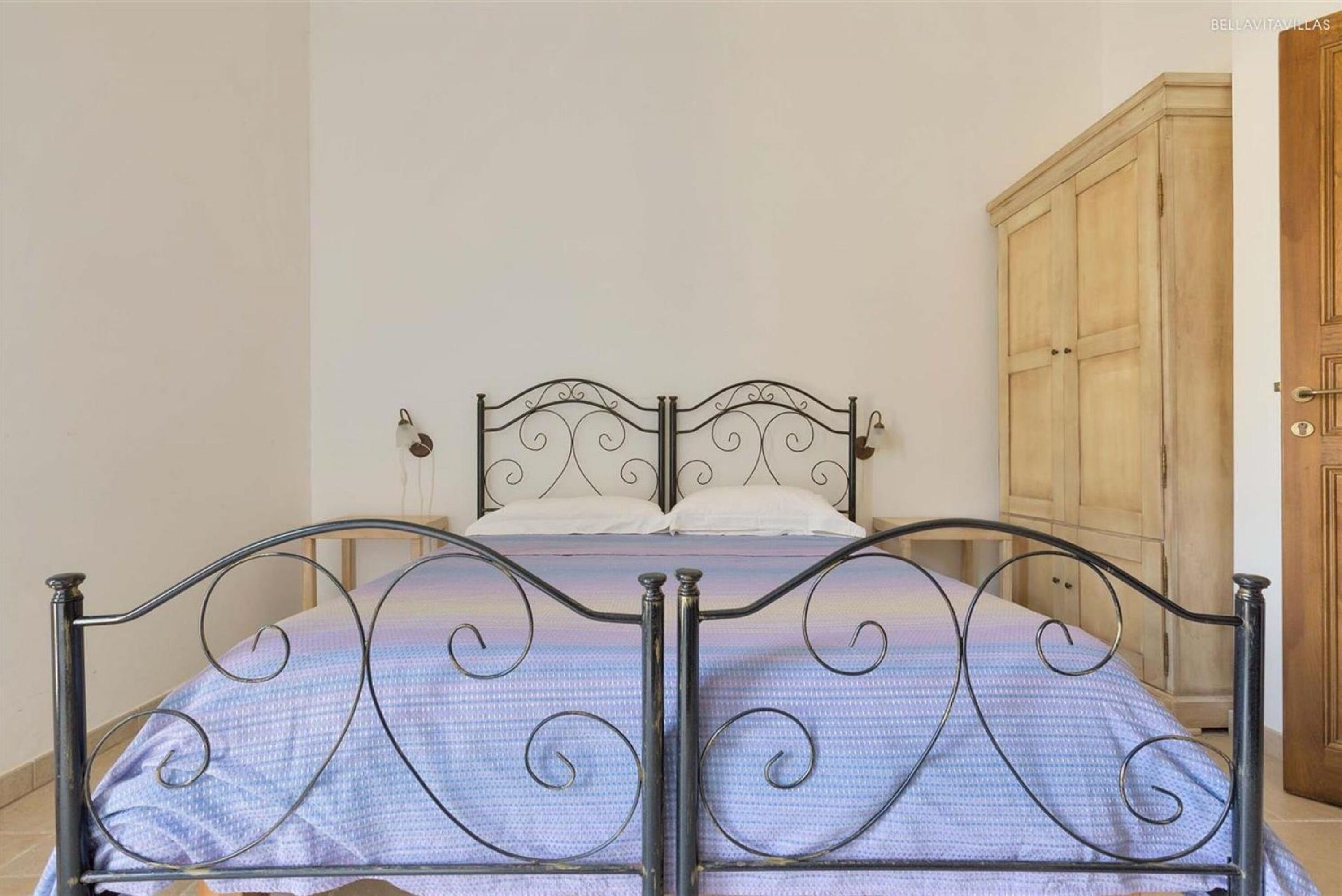 Uma cama com estrutura metálica e colcha estampada, um guarda-roupa de madeira e uma porta de madeira em um quarto com paredes lisas.