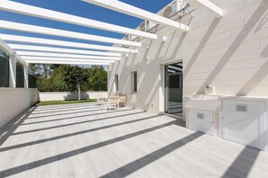 Um pátio moderno ao ar livre com uma pérgola branca, área de estar com cadeiras e um edifício com janelas e porta grandes.