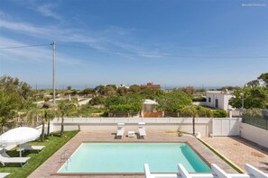 Piscina retangular com espreguiçadeiras, cerca branca e vista para o mar e edifícios ao fundo.