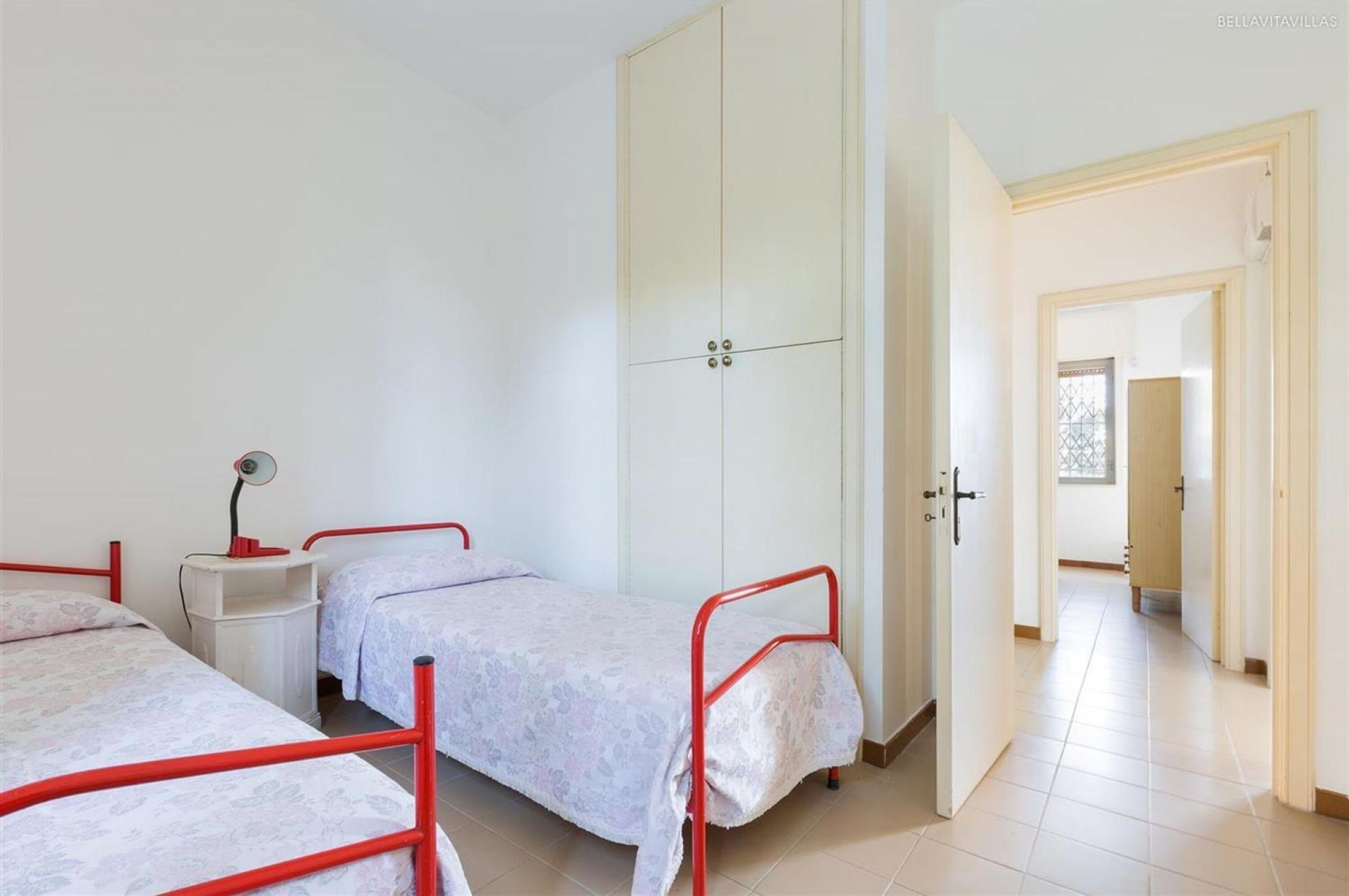 Quarto com duas camas de solteiro, guarda-roupa, mesa de cabeceira com abajur e porta que dá para outro cômodo.