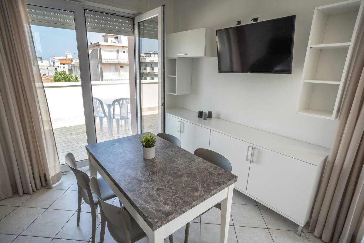 Cozinha moderna com mesa de jantar, cadeiras, televisão e varanda com vista para edifícios.