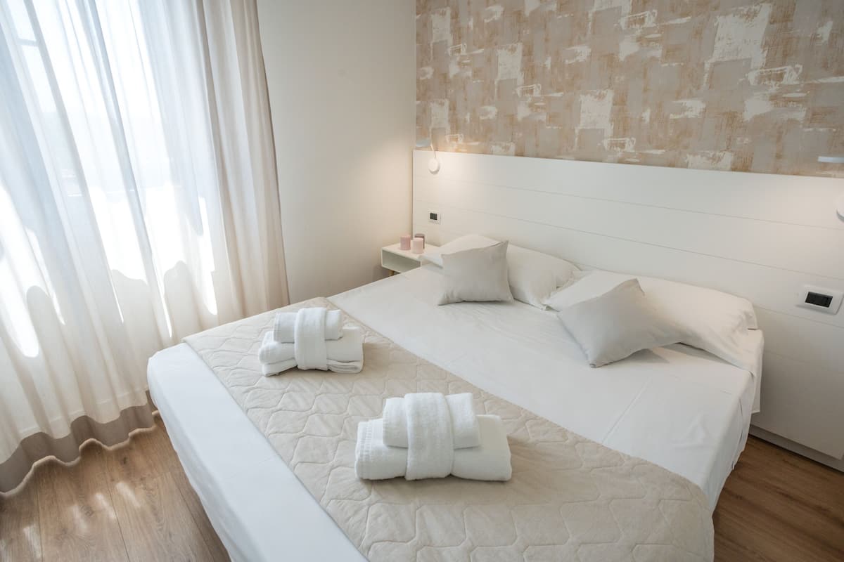 Quarto de hotel com uma cama grande, duas almofadas, uma mesa de cabeceira com luminária e uma parede com textura.