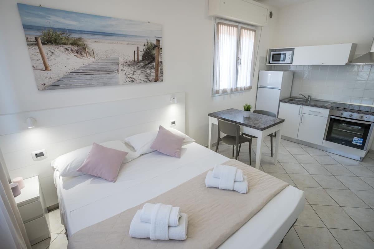 Quarto de hotel com cama, escrivaninha, cozinha compacta e pintura com vista para a praia.