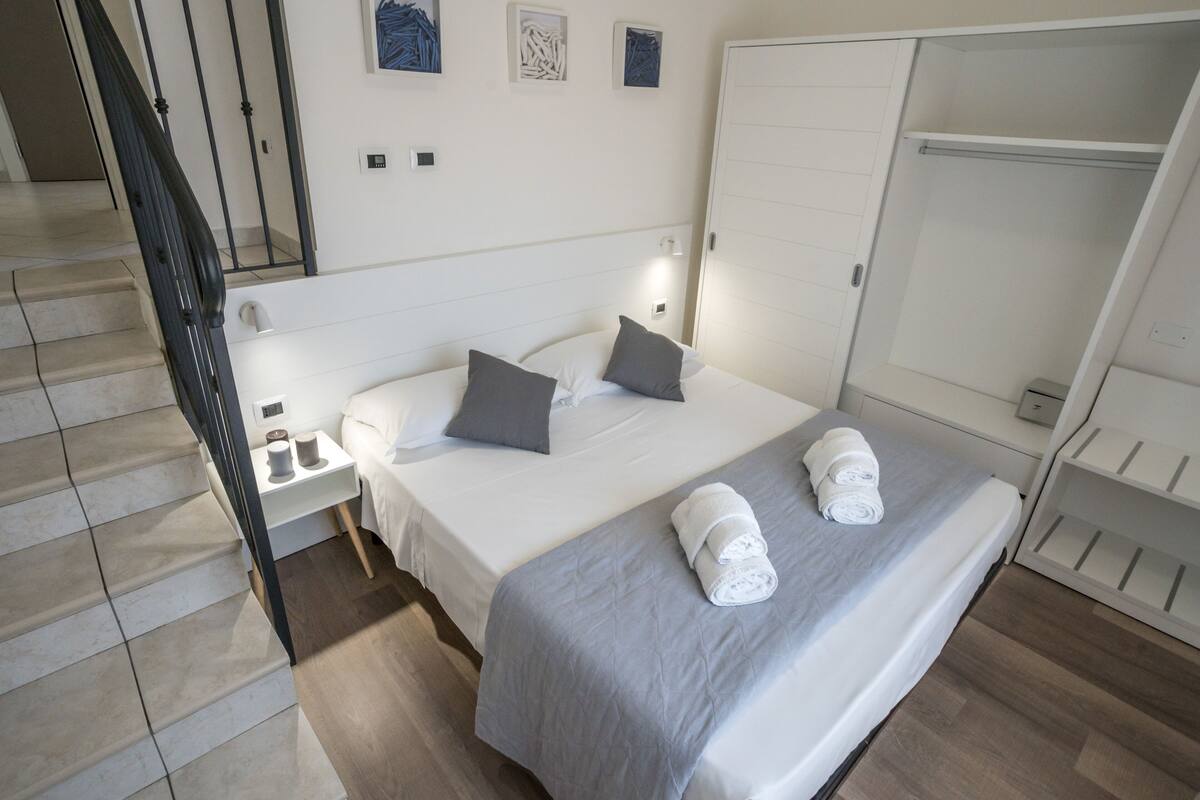 Quarto de hotel com cama, duas mesas de cabeceira, um guarda-roupa e uma escadaria.