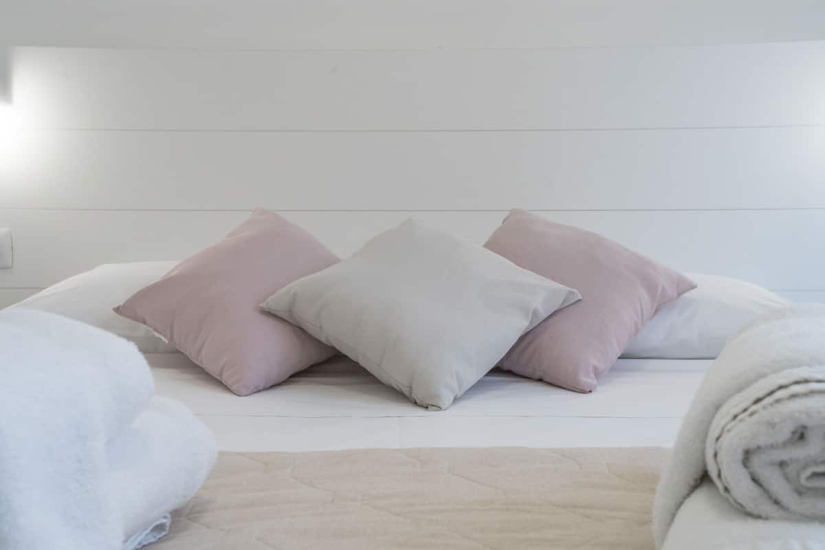 Cama bem arrumada com três almofadas em tons de rosa e branco, uma toalha branca enrolada e um tapete bege.
