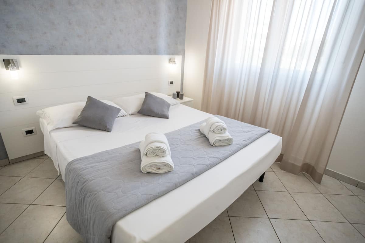 Quarto de hotel moderno com uma cama grande, roupa de cama cinza e toalhas dobradas com cuidado.