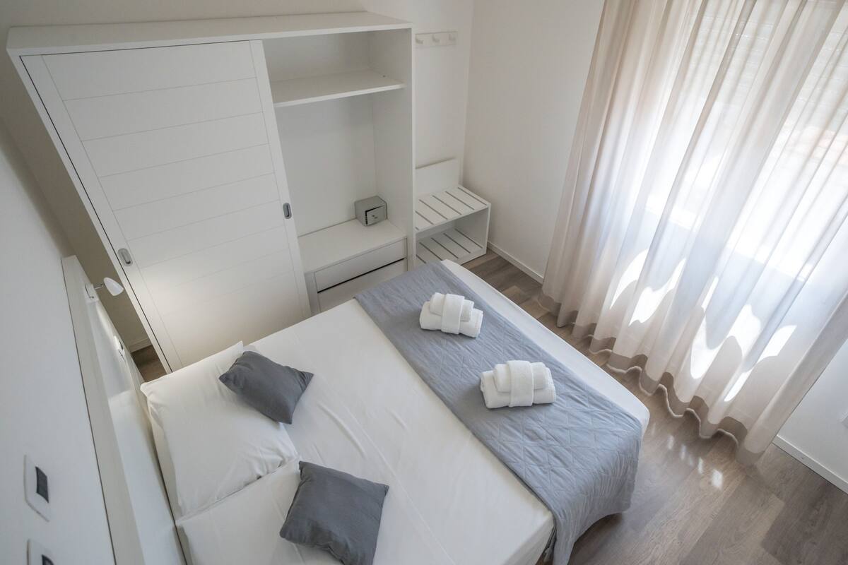 Quarto de hotel com cama, duas almofadas, mesa de cabeceira, guarda-roupa e janela com cortinas.