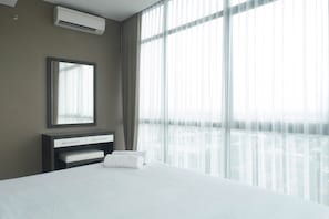 Quarto de hotel com uma cama grande, mesa de cabeceira com espelho, um banquinho e vista para a cidade através de cortinas translúcidas.