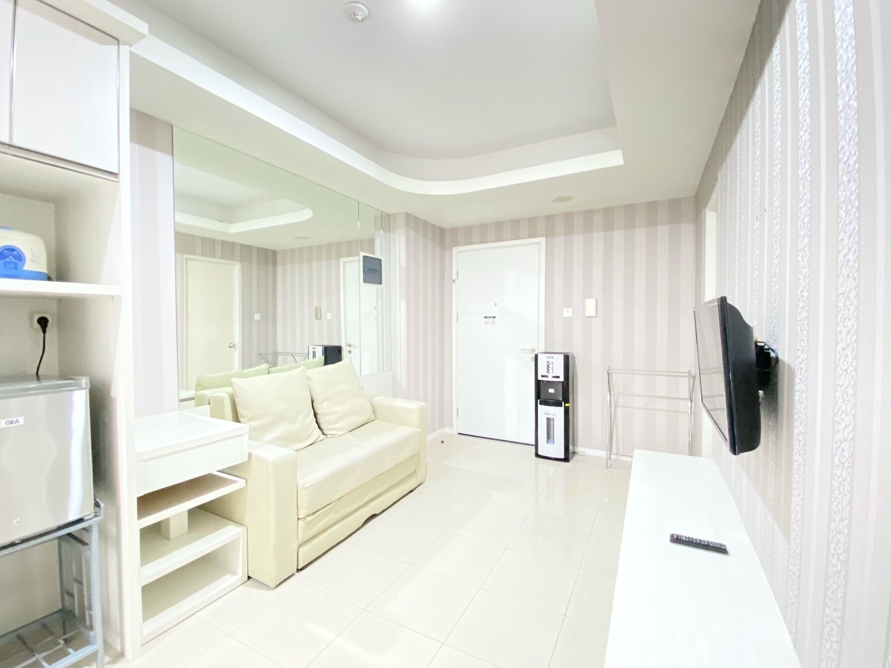 Quarto de hotel moderno com sofá, uma mesinha, televisão fixada na parede e um frigorífico.