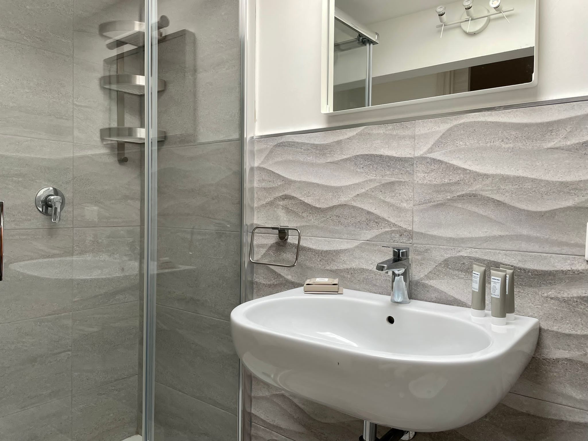 Casa de banho com cabine de duche em vidro, lavatório branco com uma torneira e parede com textura atrás do lavatório.