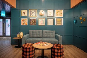 Quarto com paredes verde-água, sofá cinza, dois pufes xadrez e diversas obras de arte emolduradas.