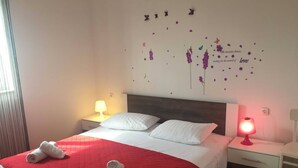 Cama bem arrumada com manta vermelha, dois travesseiros brancos e duas toalhas enroladas. Duas mesas de cabeceira com luminárias, uma de cada lado da cama. Na parede, decoração floral com flores roxas e borboletas.