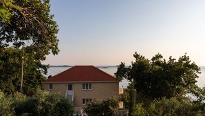 Uma casa de dois andares com telhado de telhas vermelhas, rodeada por vegetação e vista para o mar.