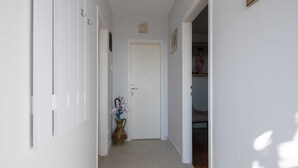 Um corredor estreito com uma porta branca no final, um vaso com flores e um pequeno objeto decorativo na parede.